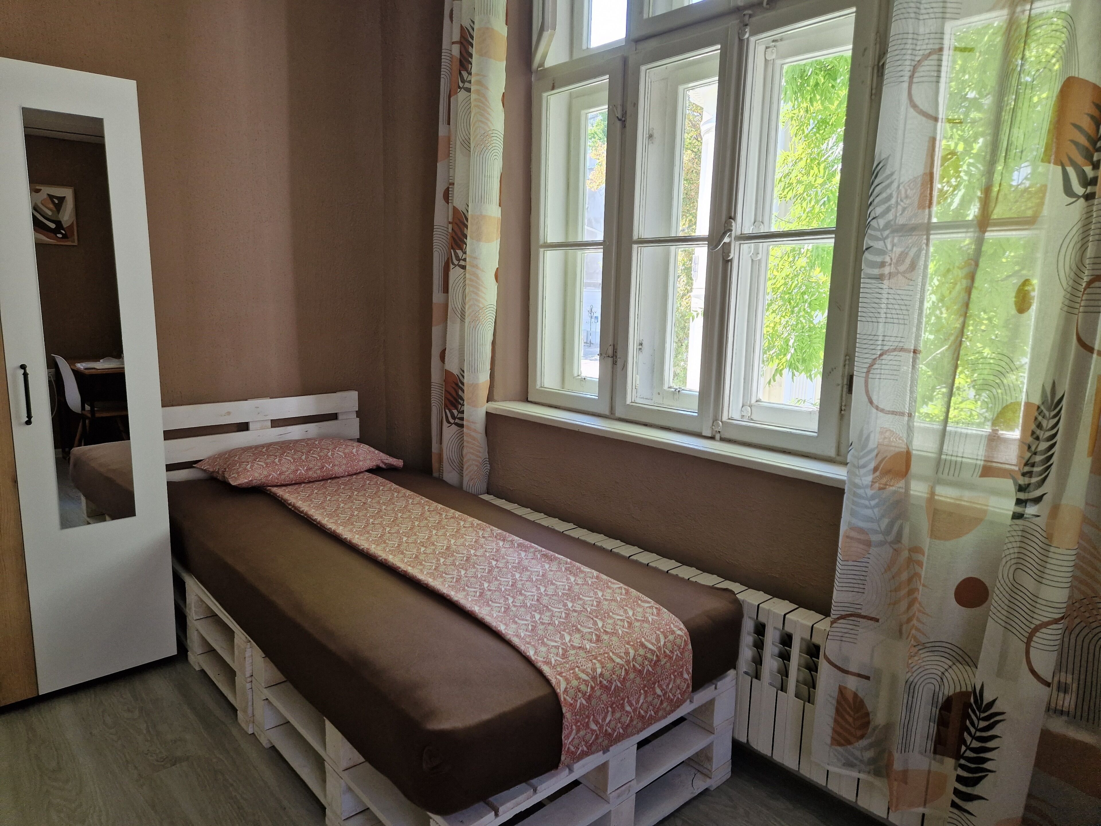 Quarto pequeno com cama de solteiro, janela, radiador e porta para outro espaço.