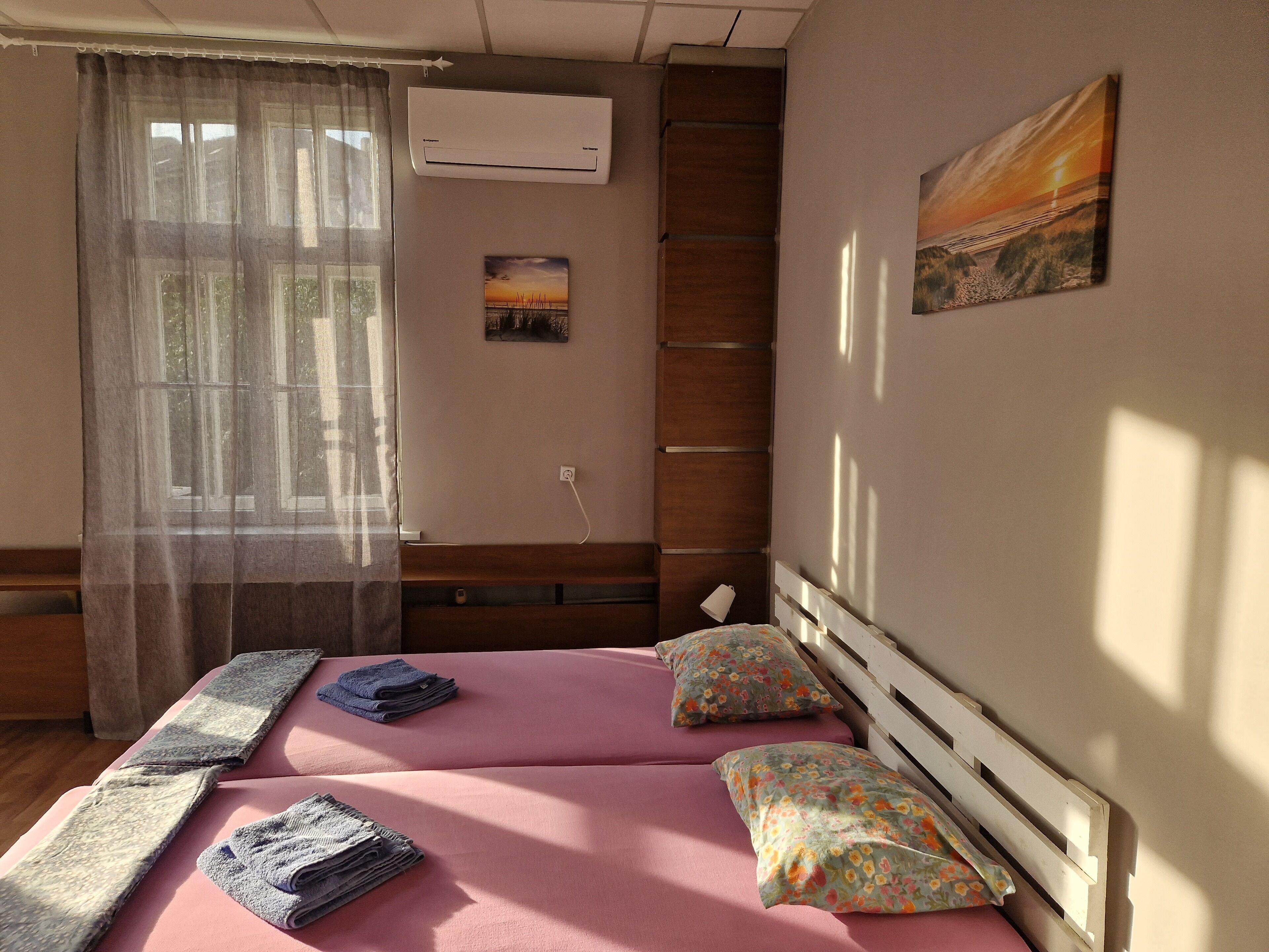 Quarto com duas camas, janela com cortinas leves, aparelho de ar condicionado, parede de painel de madeira e uma pintura de pôr do sol.