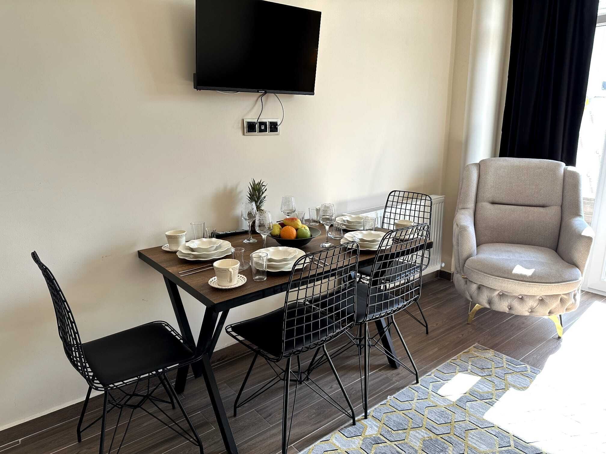 Uma sala de jantar com mesa posta para quatro pessoas, uma televisão montada acima e uma poltrona confortável.
