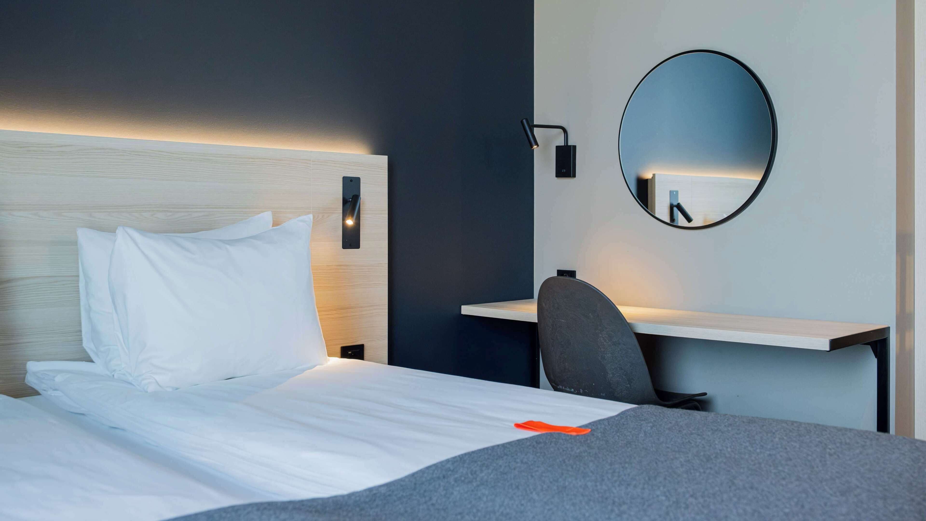 Quarto de hotel com cama, escrivaninha e espelho redondo.