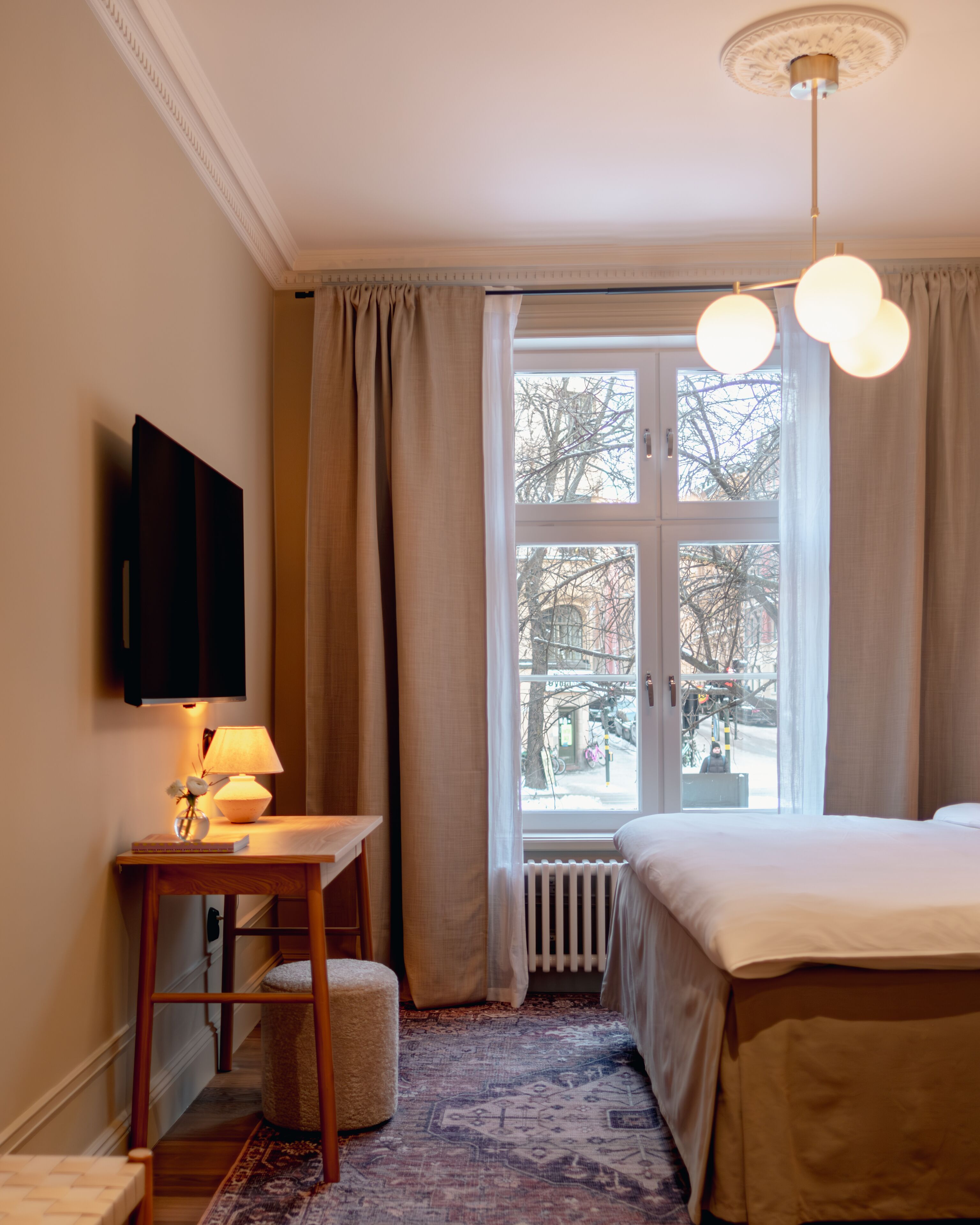 Quarto de hotel com cama, escrivaninha com abajur, televisão na parede e janela com cortinas.