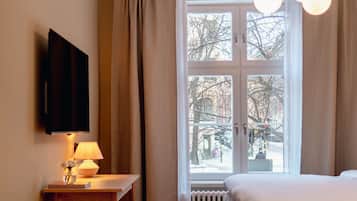 Quarto de hotel com cama, escrivaninha com abajur, televisão na parede e janela com cortinas.