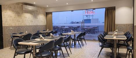 Restaurante com mesas e cadeiras dispostas para refeições, uma grande janela com vista e um aparelho de ar condicionado na parede.