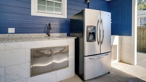 Cozinha com geladeira de aço inoxidável, janela acima da pia e parede externa azul.