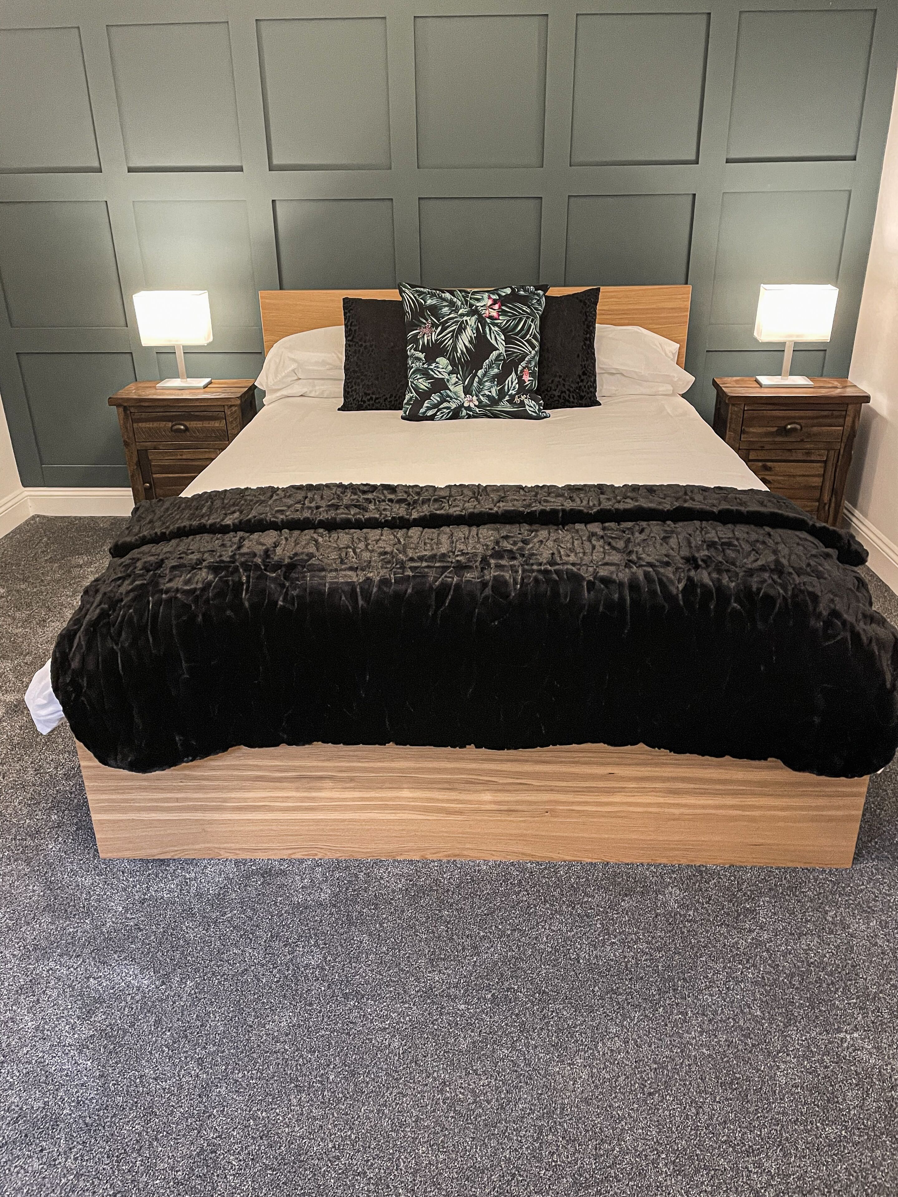 Uma cama com edredom preto, duas mesinhas de cabeceira de madeira, um travesseiro estampado com flores e uma parede com painéis.