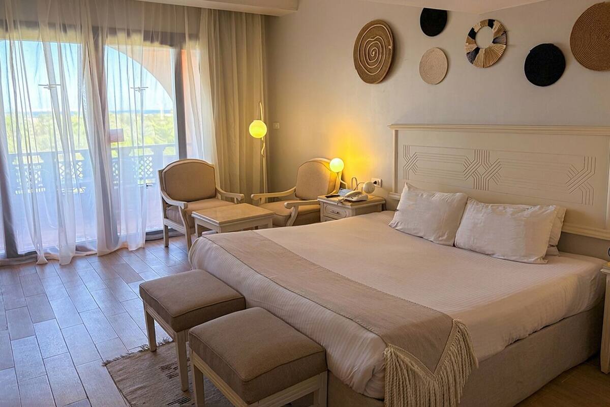 Quarto com cama grande, mesa de cabeceira, cadeira e vista pela janela com cortinas leves.