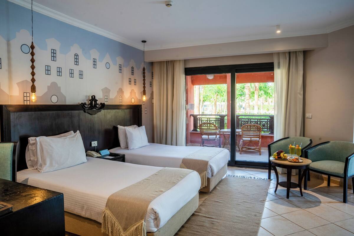 Quarto de hotel com duas camas, área de jantar com cadeiras, mesa com frutas e bebidas, e vista para o exterior.