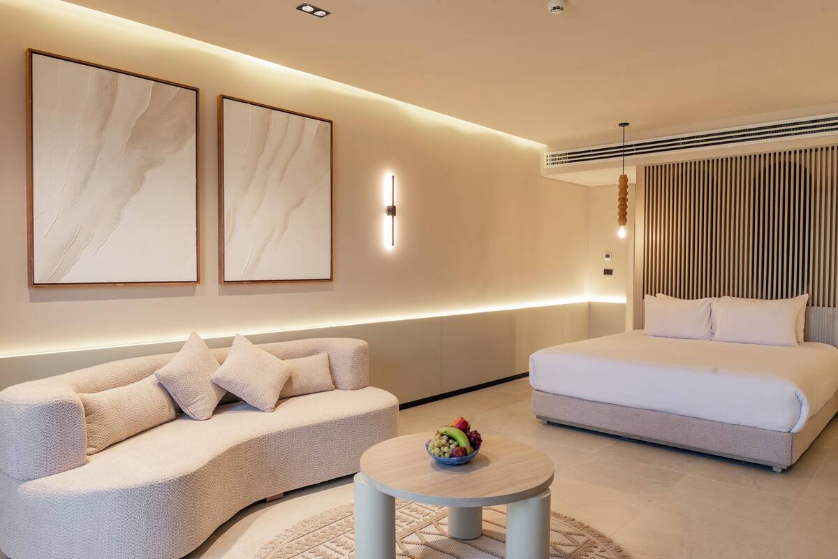 Quarto de hotel moderno com sofá, mesa redonda com frutas, cama e duas luminárias fixadas na parede.