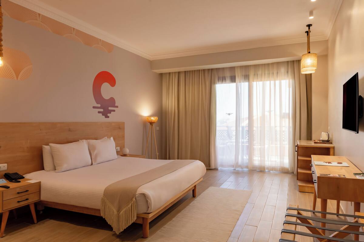 Quarto de hotel com uma cama grande, mesas de cabeceira, escrivaninha, cadeira, televisão e uma janela ampla com cortinas.