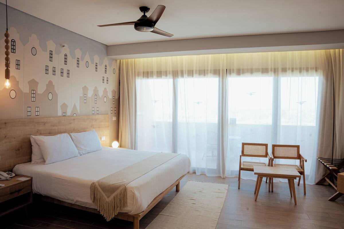 Quarto de hotel com uma cama, duas cadeiras de madeira, um ventilador de teto e uma janela grande com cortinas leves.