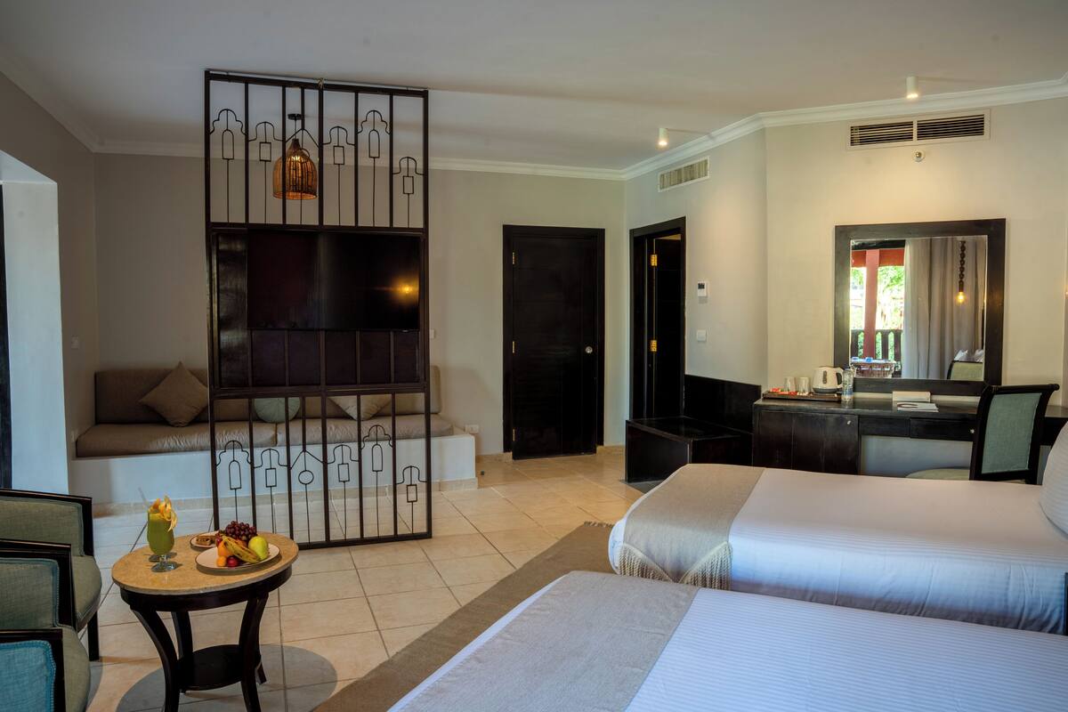 Quarto de hotel com duas camas, uma escrivaninha, televisão, sofá e mesa de jantar com frutas.