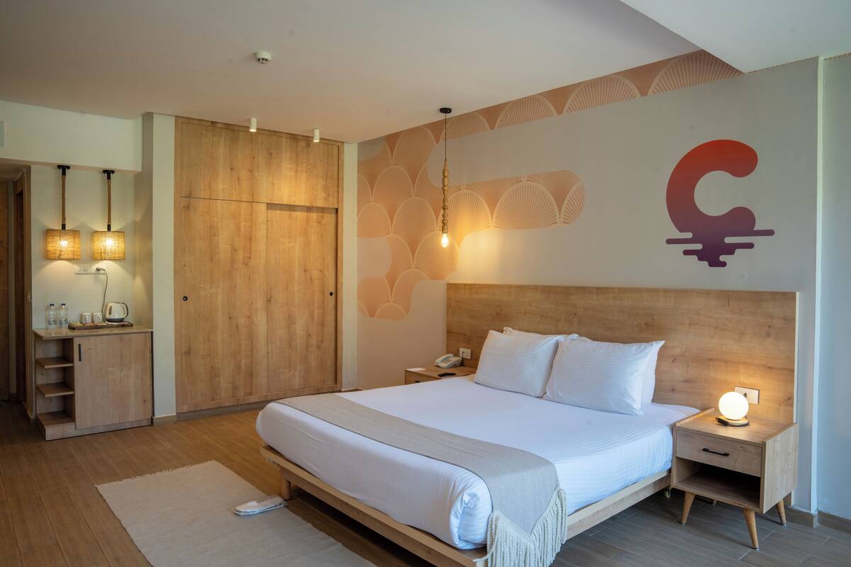 Quarto com cabeceira de madeira, mesa de cabeceira com abajur, criadeira e cama com roupa de cama branca, além de uma parede com padrão decorativo.