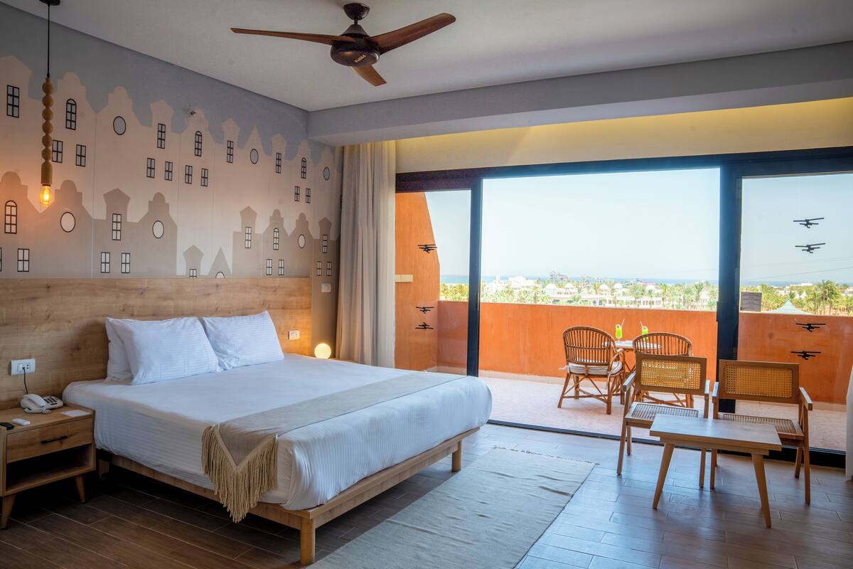 Quarto com uma cama grande, mesa de cabeceira, ventilador de teto e varanda com cadeiras e mesa.