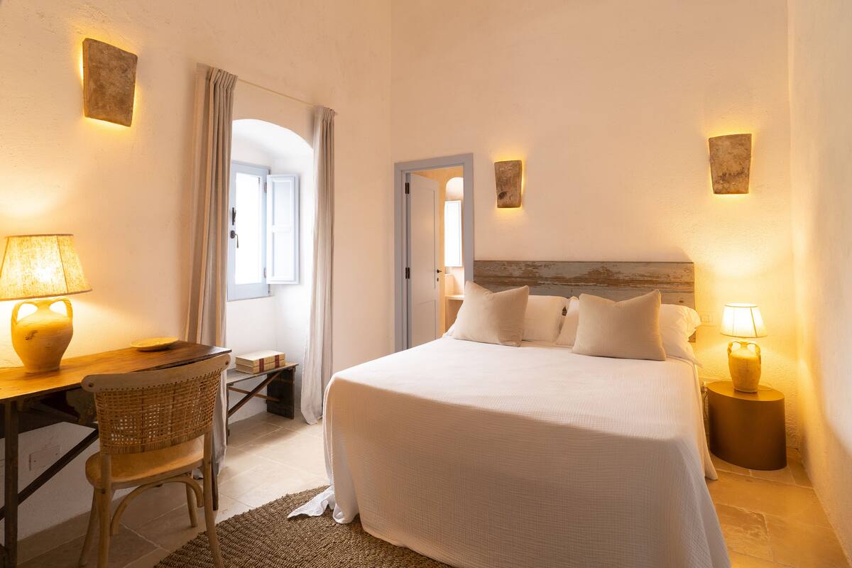 Quarto de hotel com uma cama grande, uma escrivaninha com luminária, uma cadeira e uma mesinha.