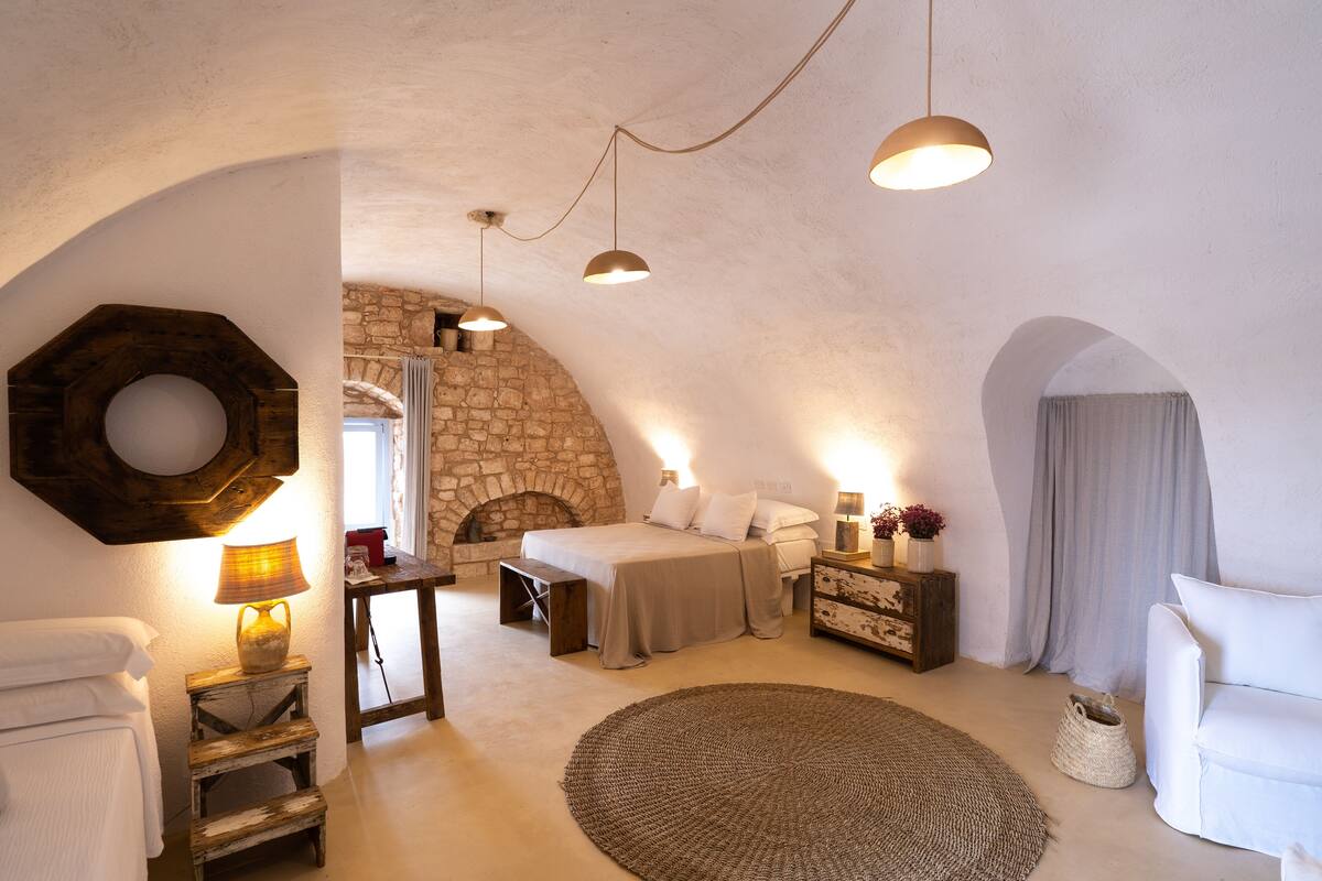 Um quarto acolhedor com lareira de pedra, uma cama com colcha cinzenta, uma mesinha de cabeceira de madeira, um sofá branco e um tapete tecido.