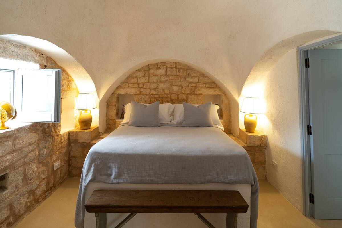 Quarto com teto abobadado em pedra, cama com roupa de cama branca, duas luminárias de mesa e um banco de madeira.