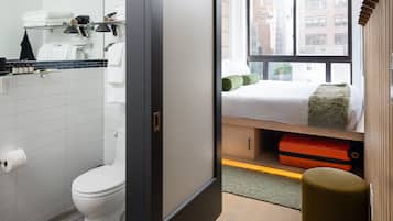 Um banheiro moderno com vaso sanitário, pia e espelho. No quarto vizinho, há uma cama com travesseiro verde e cobertor branco.