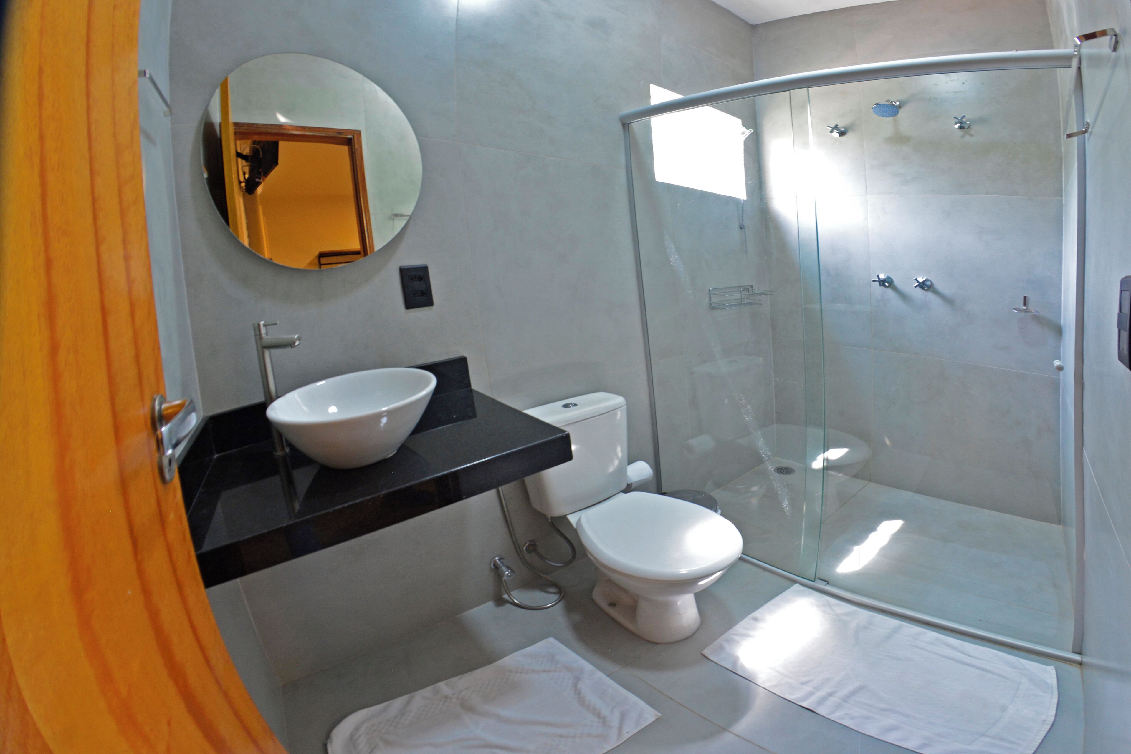 Uma casa de banho moderna com cabine de duche de vidro, espelho redondo, lavatório branco e sanita.