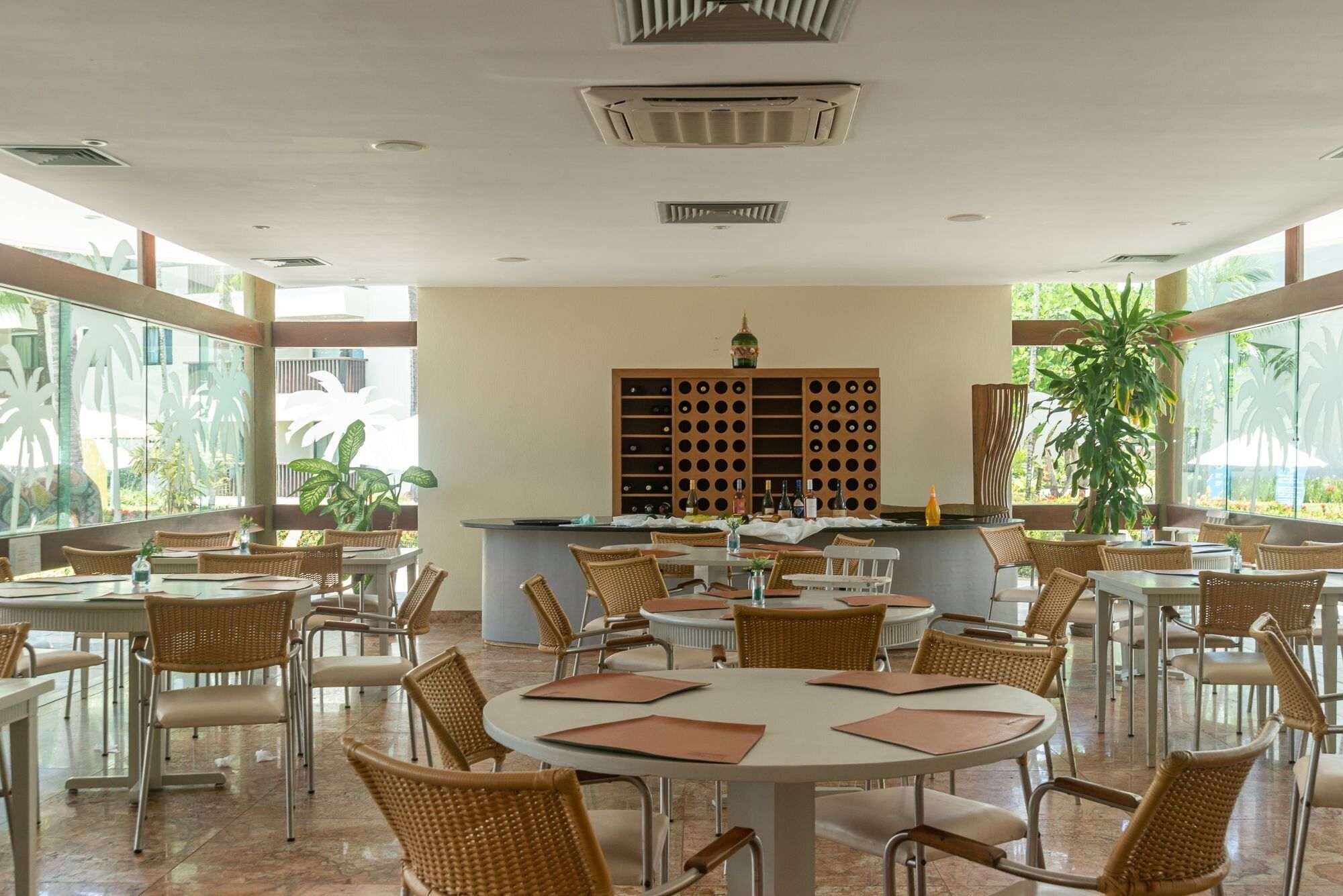Restaurante com mesas redondas e cadeiras de vime, adega e janelas amplas com vista para palmeiras.