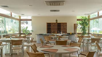 Restaurante com mesas redondas e cadeiras de vime, adega e janelas amplas com vista para palmeiras.