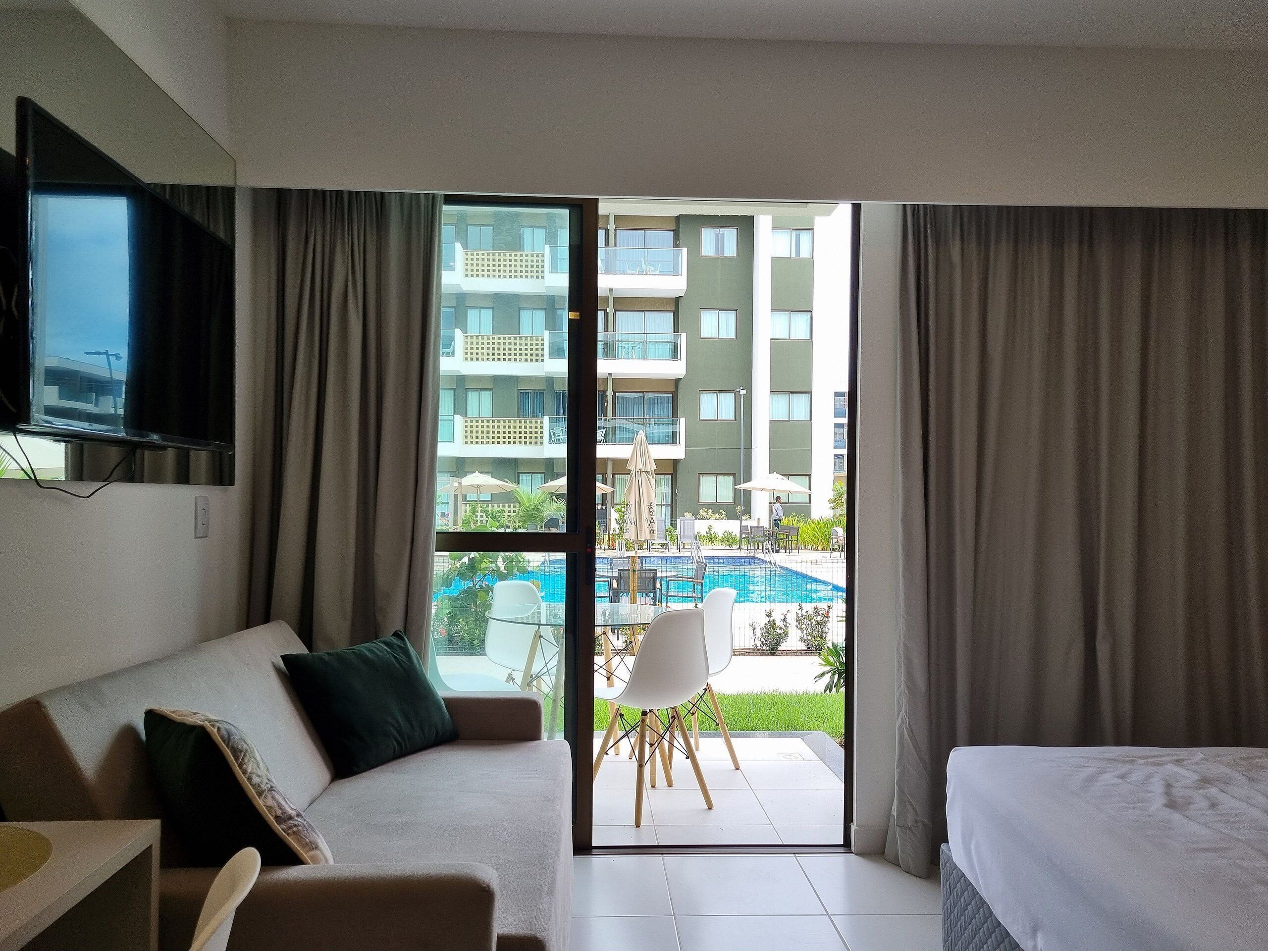 Quarto de hotel com sofá, cama, televisão na parede e vista para uma piscina ao ar livre e um edifício.