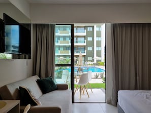Quarto de hotel com sofá, cama, TV fixada na parede e vista para uma piscina e um edifício.