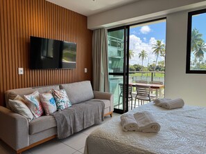 Quarto de hotel moderno com sofá, cama com toalhas dobradas, televisão de tela plana fixada em parede de madeira e varanda com vista para palmeiras.