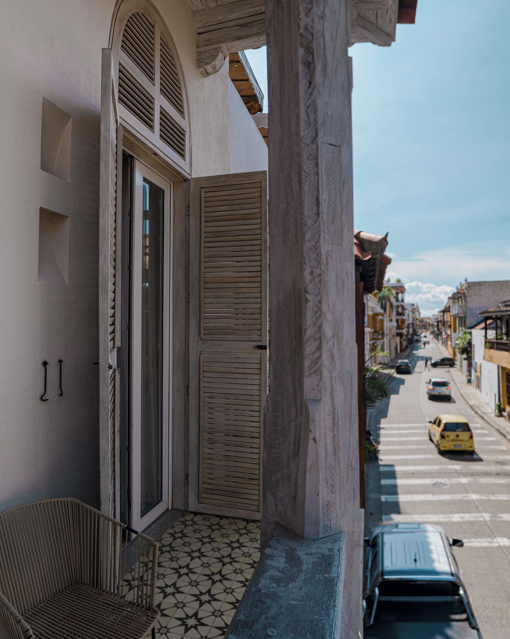 Varanda com cadeira de vime, porta fechada e vista para a rua com carros e prédios.
