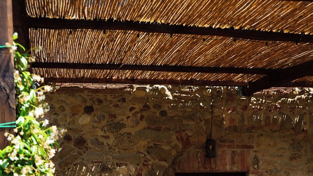 Um quarto com paredes de pedra, uma janela grande, uma cadeira de vime e uma mesa com toalha branca.