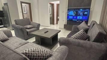 Uma sala de estar moderna com sofás cinza, uma mesa de centro e uma TV de tela plana exibindo diversos canais.