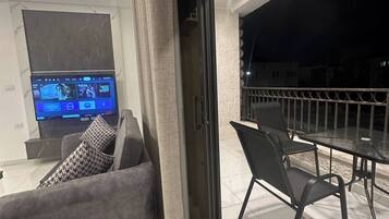 Uma sala de estar moderna com um sofá, uma televisão e uma porta de vidro que dá para uma varanda externa com mesa e cadeiras.