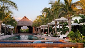 Uma área de piscina com espreguiçadeiras e guarda-sóis, cercada por palmeiras e uma estrutura com telhado de palha.