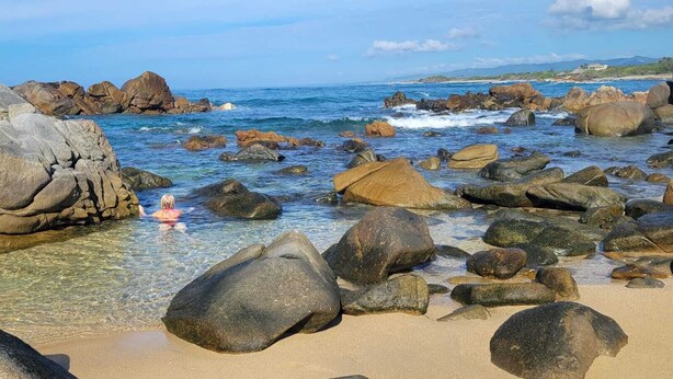 Uma praia rochosa com água cristalina, pedras grandes e uma pessoa nadando.