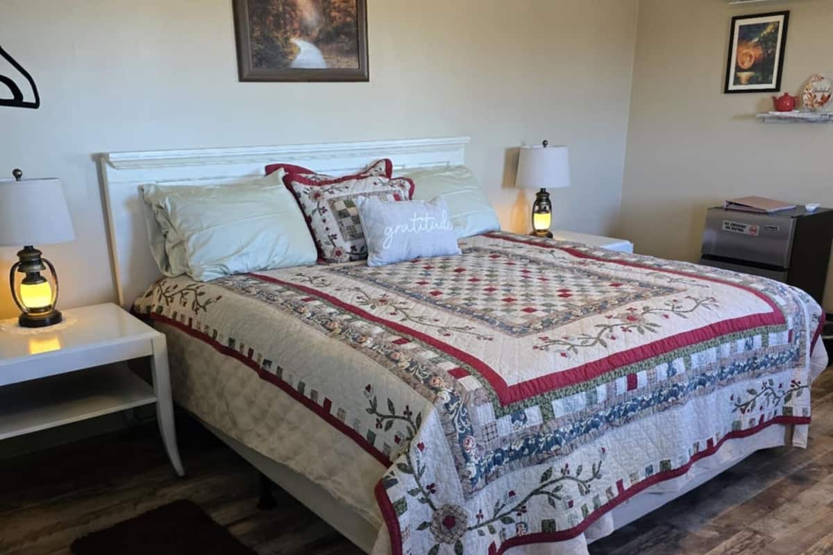Uma cama bem arrumada com um edredom floral, duas luminárias de cabeceira, um criado-mudo, um tapete pequeno e um ventilador de teto.