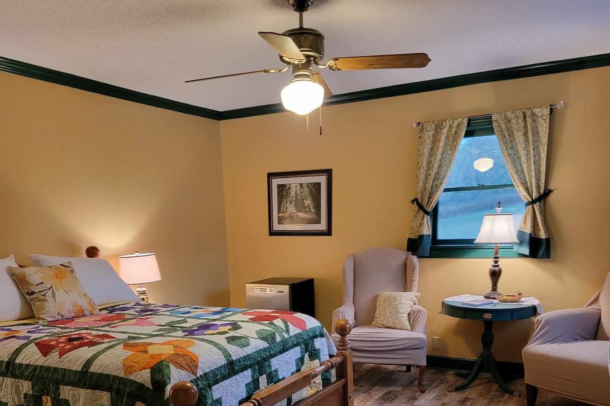 Um quarto com cama de madeira, ventilador de teto, janela com cortinas, uma cadeira e uma mesa pequena com abajur.