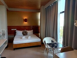 Quarto de hotel com cama, mesa de cabeceira, cadeira e uma pequena mesa com um ventilador.