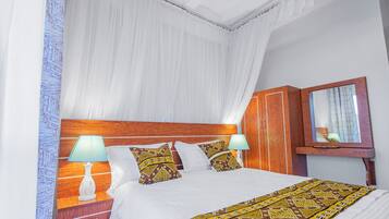 Quarto com cama de dossel, cabeceira de madeira, luminárias de cabeceira, almofadas estampadas e mesa de cabeceira.