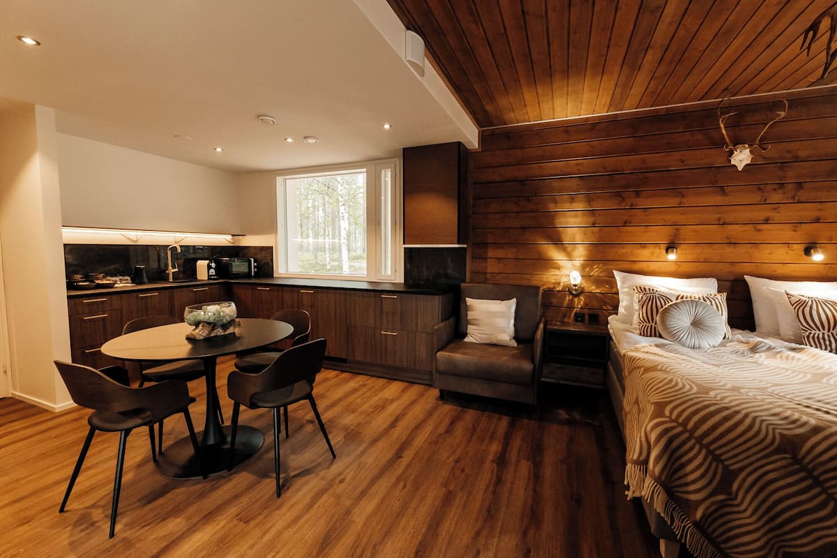 Quarto compacto com cozinha, mesa de jantar com cadeiras, sofá e cama com roupa de cama estampada.