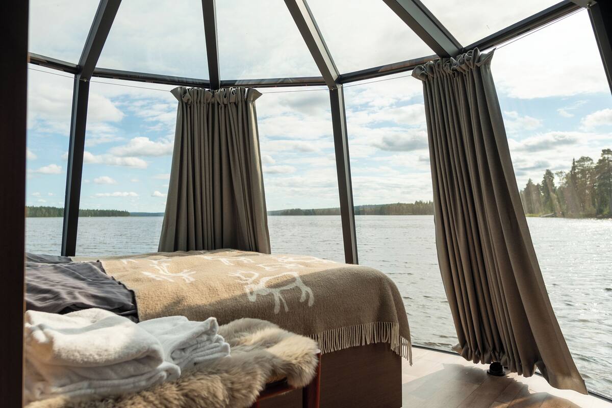 Quarto com cama, manta de pele e vista para um lago através de amplas janelas.