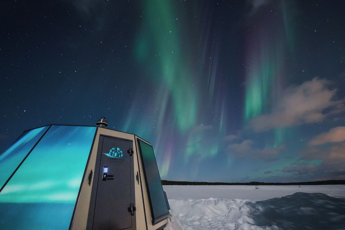 Paisagem coberta de neve com um edifício que possui uma grande janela e uma porta, sob um céu noturno com estrelas visíveis e aurora boreal.