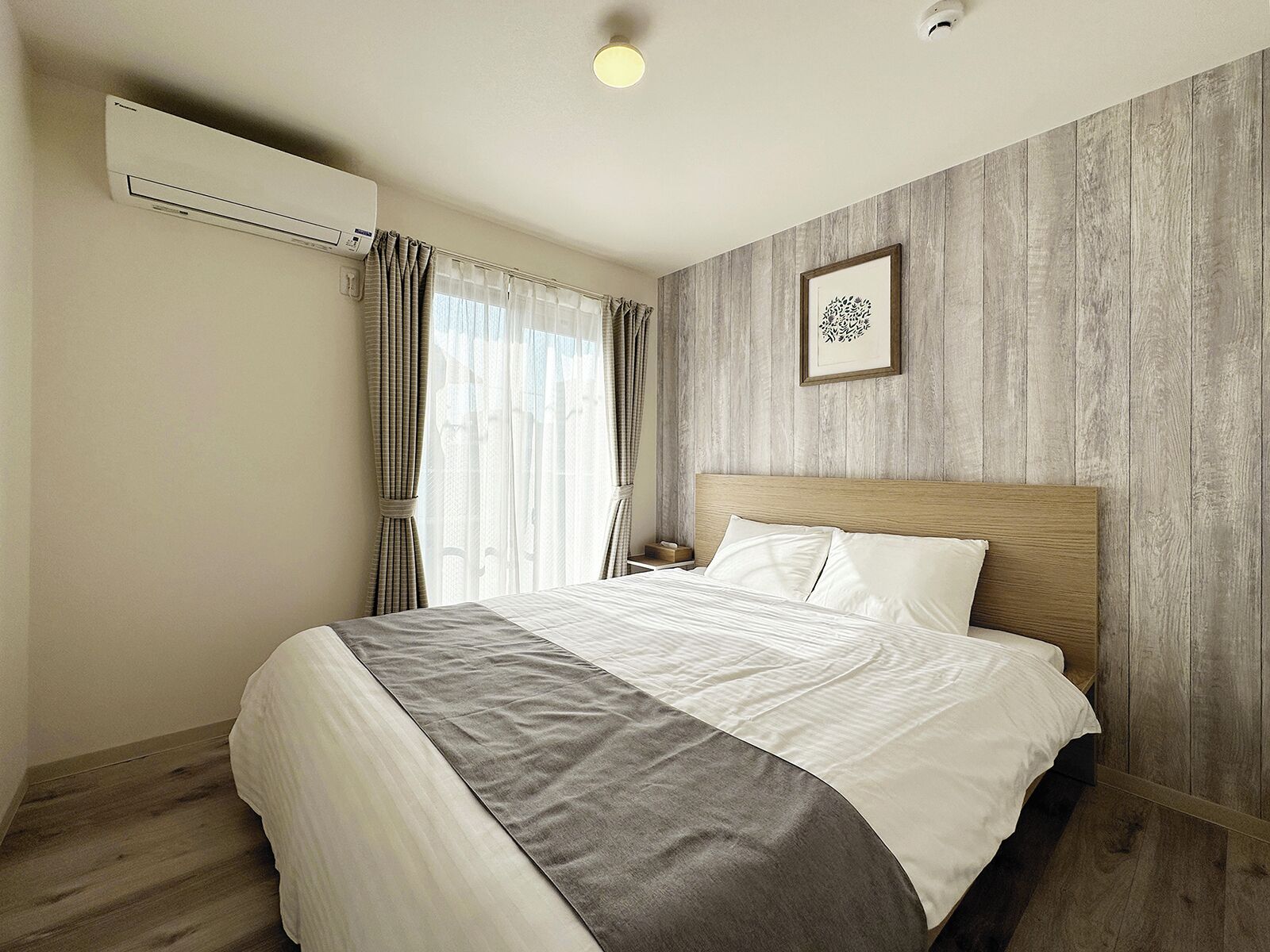 Quarto de hotel com cabeceira de madeira, cama com roupa de cama branca e cinza, janela com cortinas leves e um ar condicionado fixado na parede.