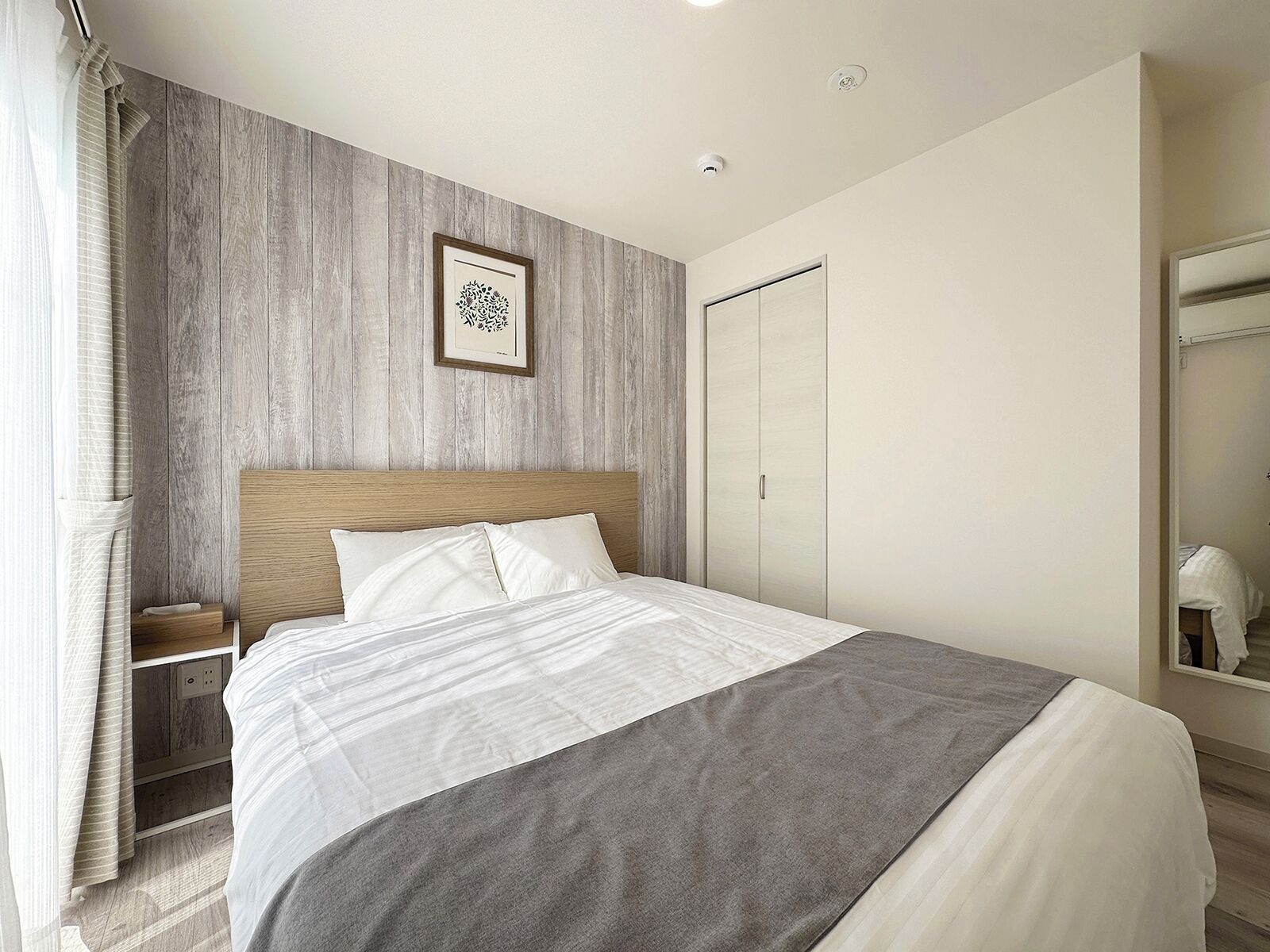 Quarto moderno com cabeceira de madeira, colcha cinza, mesa de cabeceira, guarda-roupa e uma obra de arte emoldurada na parede.