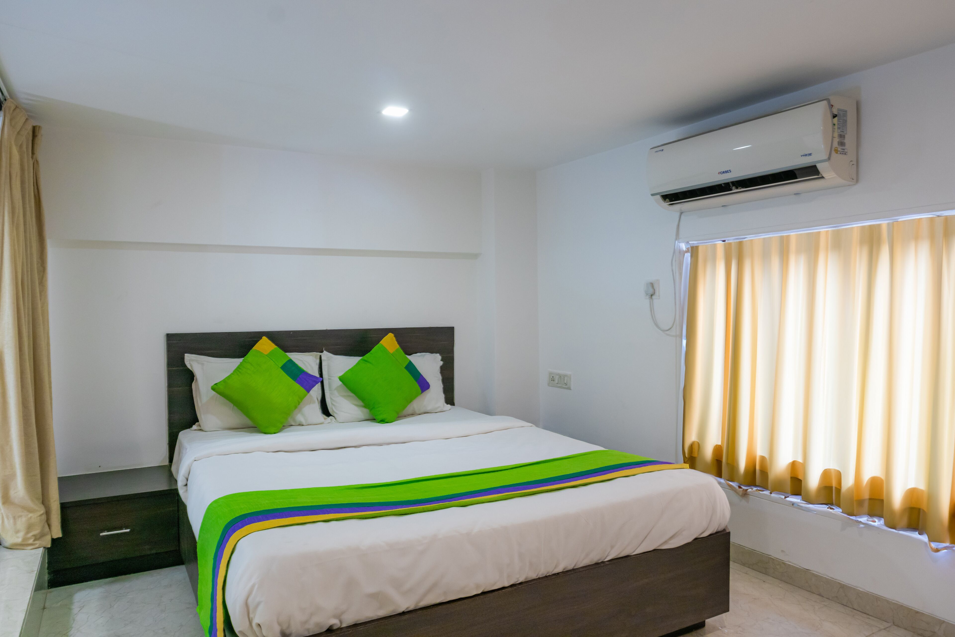 Quarto de hotel com cama, mesa de cabeceira, janela com cortinas, aparelho de ar condicionado e tomada na parede.
