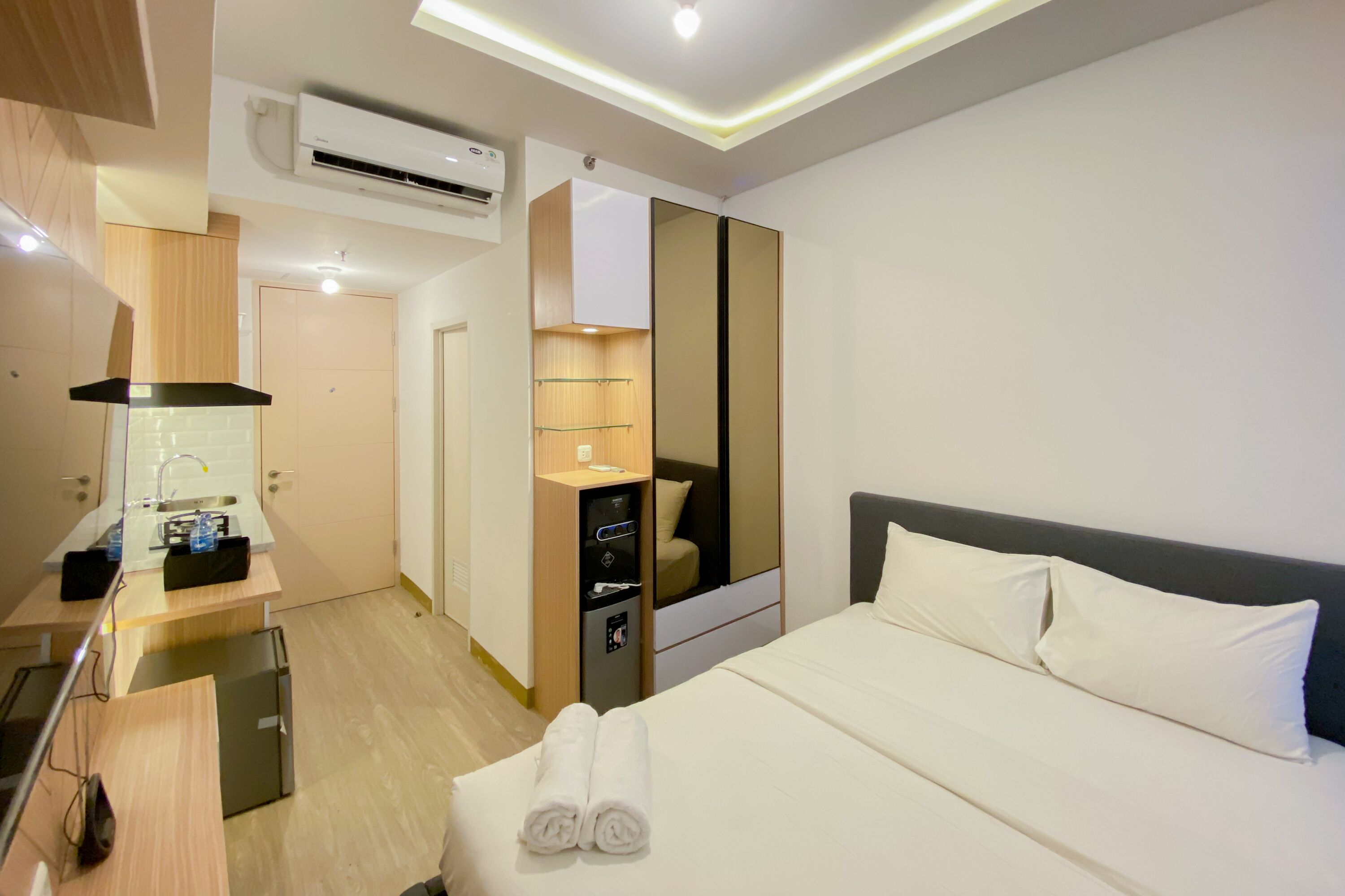 Quarto de hotel moderno com cama, escrivaninha, mini-geladeira, espelho e armário embutido.