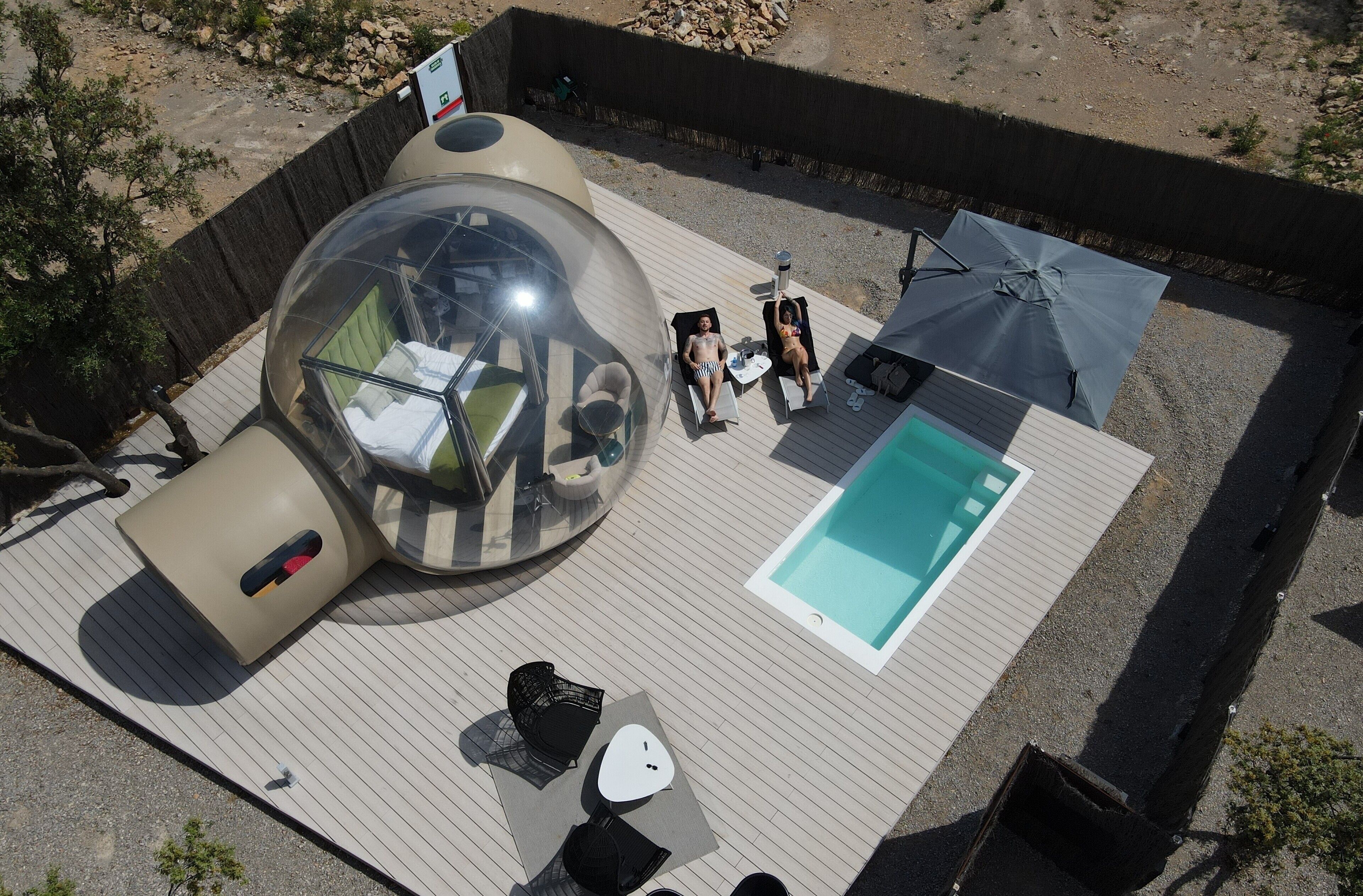 Uma sala de vidro esférica com piscina e espreguiçadeiras em um deck de madeira.