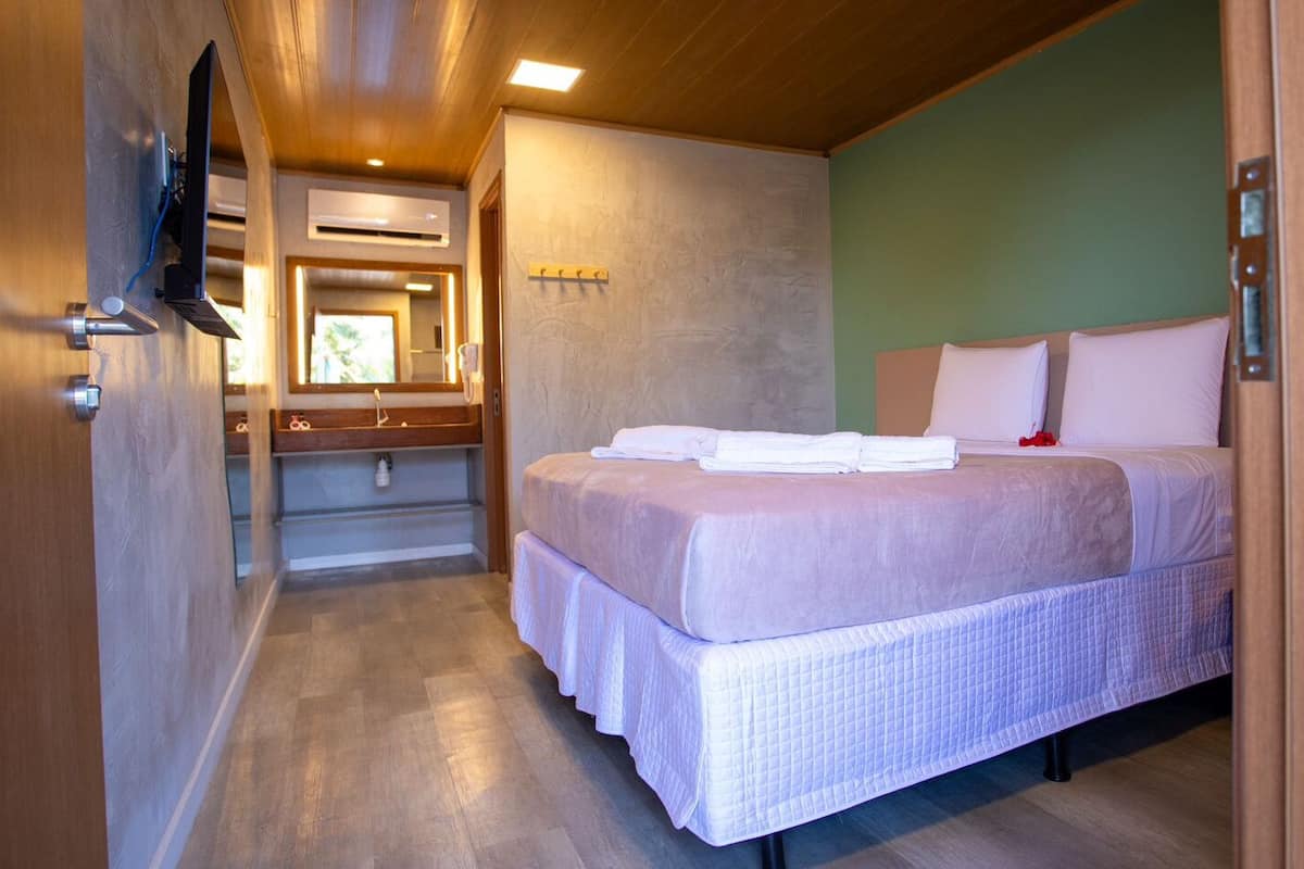 Quarto compacto com cama, mesa de cabeceira e televisão na parede.