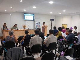Uma sala de conferências com um palestrante no púlpito, participantes sentados em mesas e uma tela de projeção.