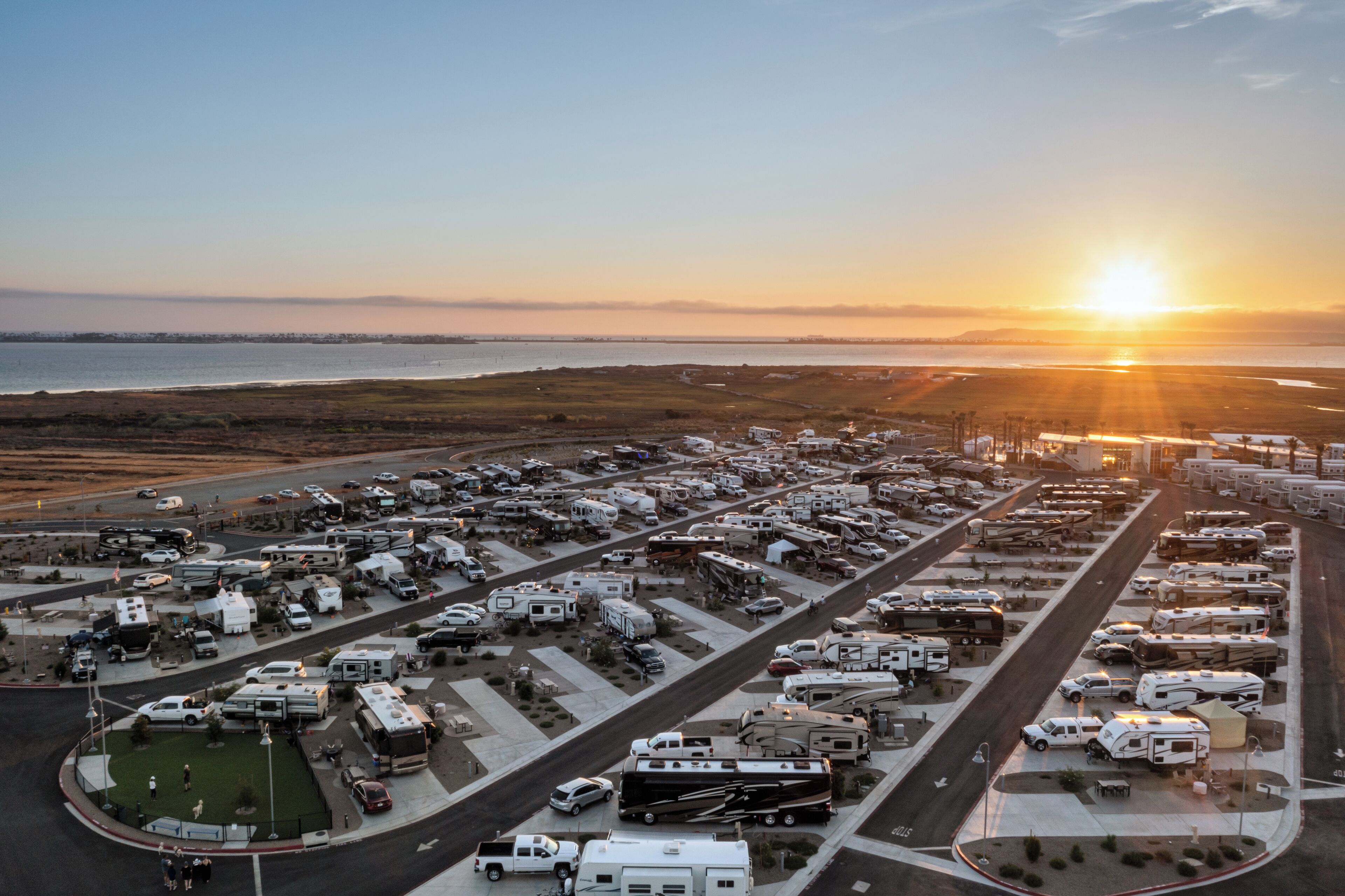 Um parque para veículos recreativos com várias fileiras de RVs e trailers, uma estrada e um pôr do sol no horizonte.