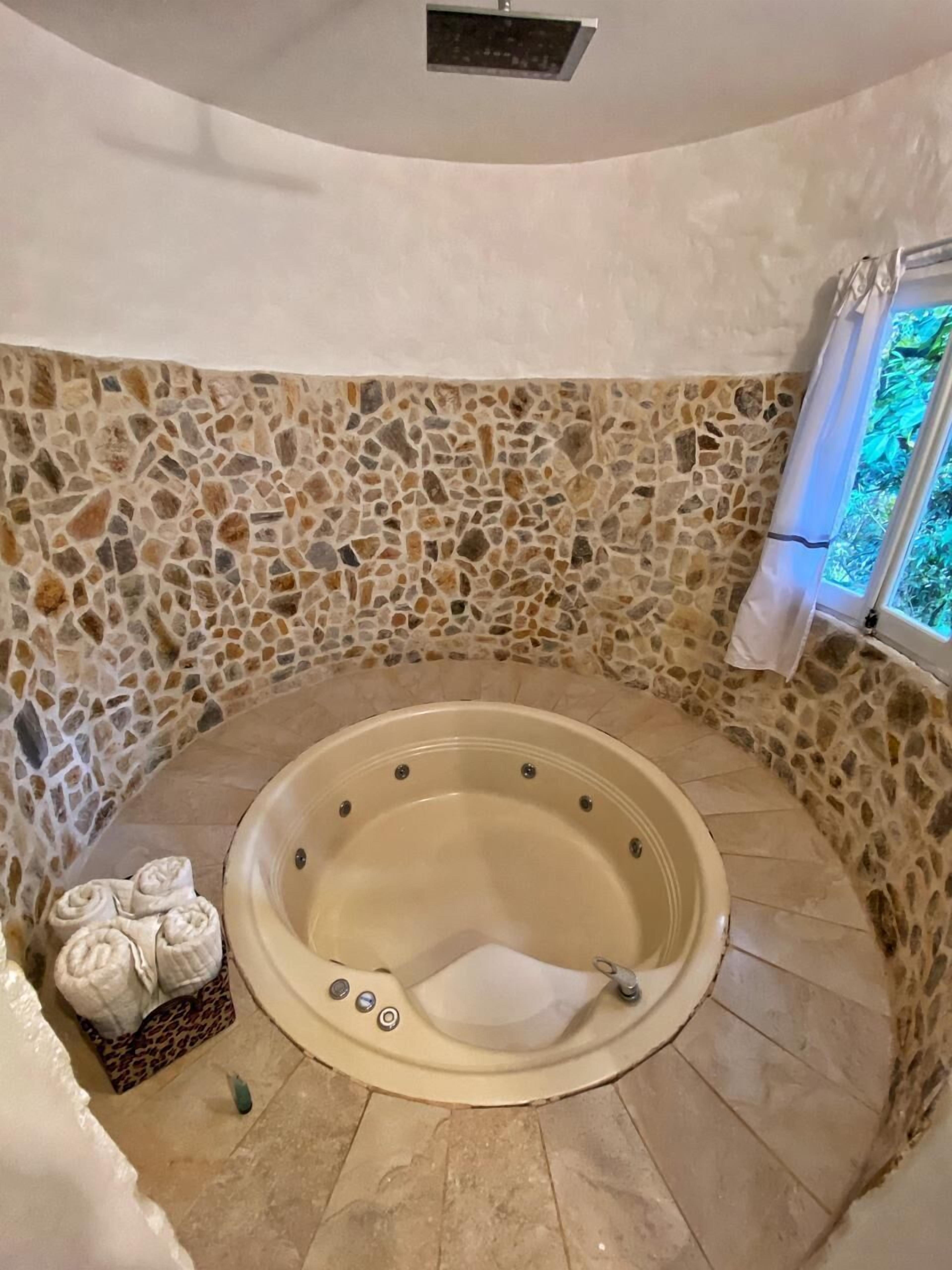 Uma banheira de hidromassagem circular com paredes em mosaico de pedra, uma janela com vista e um armário para toalhas.
