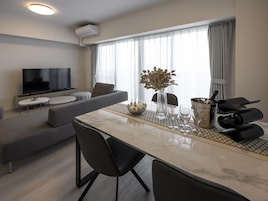 Uma sala de estar moderna com televisão de tela plana, sofá cinza e um espaço de jantar com mesa de mármore e cadeiras pretas.